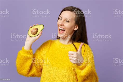 모피 스웨터를 보여주는 쾌활 한 젊은 여성은 보라색 파스텔 배경에 고립 된 신선한 잘 익은 녹색 아보카도의 절반을 들고 사람들은