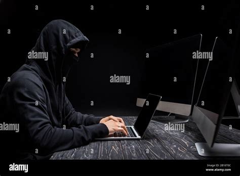 Back View Of Hooded Hacker With Hands Behind Head Near Computer Monitors With Blank Screen