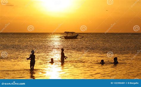 Дети играя на пляже с шлюпкой рыболова в Бали Индонезии во время захода солнца на пляже
