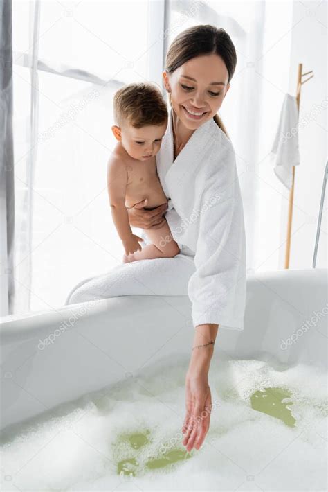 madre satisfecha y tatuada en albornoz sosteniendo en brazos al niño desnudo y llegando al agua
