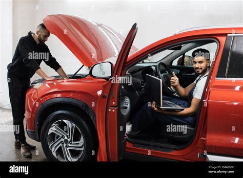 Young male mechanic using laptop sitting inside red car and his colleague with tablet under hood ...