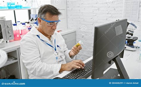 Mature Man In Lab Coat Examines Medication Bottle In Bright Laboratory While Using Computer