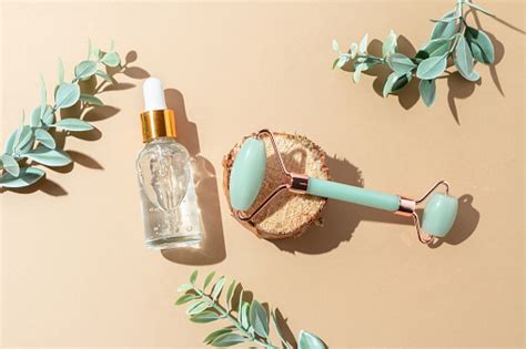 녹색 가지가있는 밝은 갈색 배경에 나무 스탠드에 점적기와 돌 구아슈 얼굴 마사지기가있는 투명한 유리 화장품 병 천연 유기농