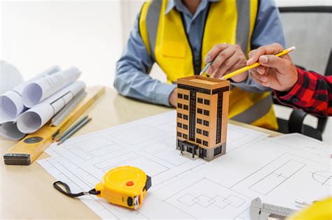 아시아계 남성 건설 계약자 엔지니어를 포함한 두 명의 남성이 건물 타워 모형이 있는 책상에서 새 집의 구조 계획과 콘크리트 기둥과 보를 엔지니어링하는 것에 대해 논의하고