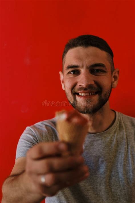 A Young Caucasian Bearded Guy Eats A Popsicle In A Cone Standing Near A Red Shabby Wall In The