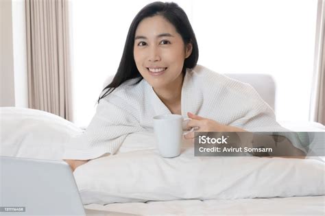 침대에 노트북에서 작업 목욕 가운에 행복 아름다운 아시아 여성 여자는 침실에서 커피 한 잔과 함께 침대에 누워 침대에 어린 소녀는 노트북 컴퓨터를 사용하고 커피를 마십니다