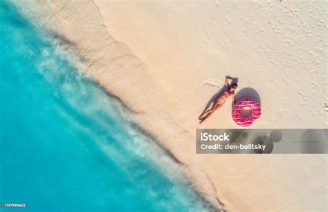 일몰에 파도와 함께 바다 근처 하얀 모래 해변에서 분홍색 도넛 수영 반지와 아름 다운 젊은 거짓말 여자의 조감도 여름 휴가 슬림 여자의 평면도 푸른 물 맑은 인도양 공중