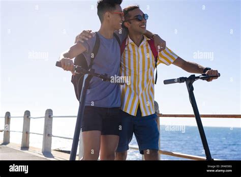 Happy Biracial Gay Male Couple Walking With Scooters Embracing On