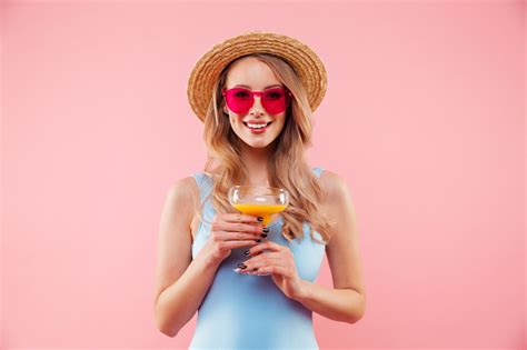 분홍색 배경에 고립 된 오렌지 주스 한 잔을 들고 미소 짓는 원피스 수영복과 밀짚 모자를 쓴 선글라스를 입은 멋진 여성