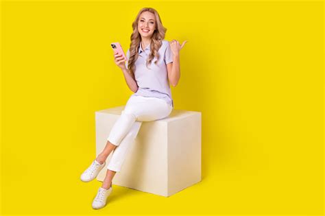 쾌활한 좋은 여자의 사진 보라색 티셔츠를 입고 스마트 폰으로 큐브에 앉아 노란색 배경에 고립 된 빈 공간을 지시 Brand Name Smart Phone에 대한 스톡 사진