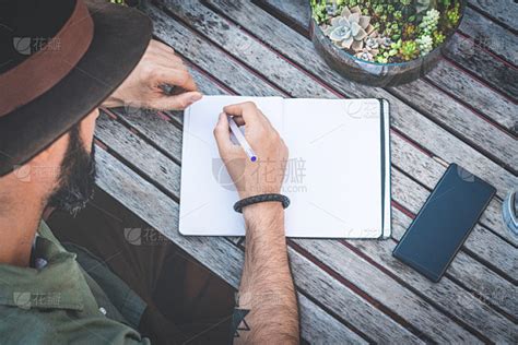 潮男学生做笔记，在笔记本上写下目标，坐在咖啡馆的露台上。聚精会神的人在木桌上写留言。前视图。