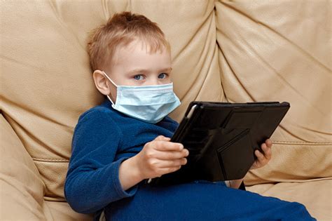 의료 용 마스크를 쓴 귀여운 아기 소년은 태블릿 패드를 손에 들고 가죽 소파에 앉아 있습니다 파란 잠옷을 입고 감성적인 얼굴 표정을 4 5세에 대한 스톡 사진 및 기타