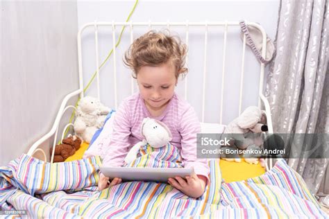 백인 5세 인 한 아이가 아기 침대에 앉아 태블릿을 손에 들고 있습니다 부드러운 장난감을 가진 곱슬 머리를 가진 소녀가 태블릿에서 재생됩니다 4 5세에 대한 스톡 사진 및