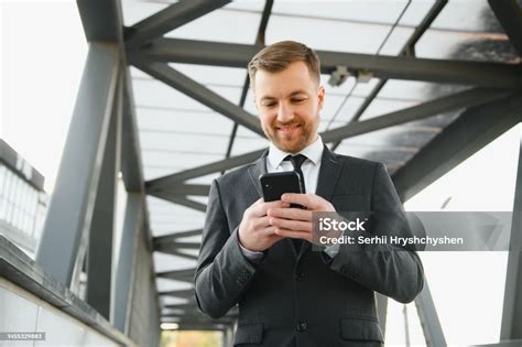 검은 양복을 입고 이른 아침에 사무실 근처에서 현대적인 스마트폰을 사용하는 행복한 미소 짓는 사업가 밤에 고층 빌딩 사무실 근처에 서 있는 동안 거래를 하는 성공적인 고용주