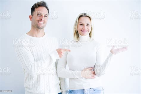 고립 된 흰색 배경 위에 서있는 캐주얼 한 티셔츠를 입은 젊은 아름다운 부부는 손으로 손가락으로 가리키는 동안 카메라에 놀랍고 미소를 지었습니다 2명에 대한 스톡 사진 및