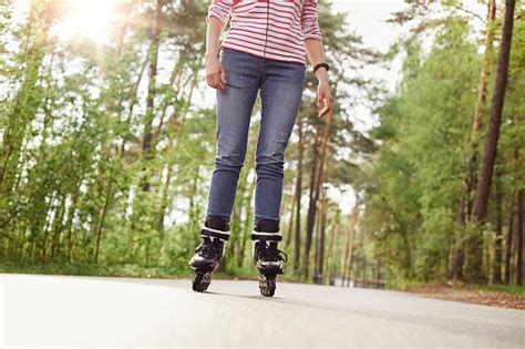 청바지와 줄무늬 셔츠를 입은 슬림한 여성의 얼굴없는 초상화 나무로 둘러싸인 아스팔트 도로에서 롤러 블레이드 활동적인 방법으로 숲에서 시간을 보내고 알 수없는 사람 롤러