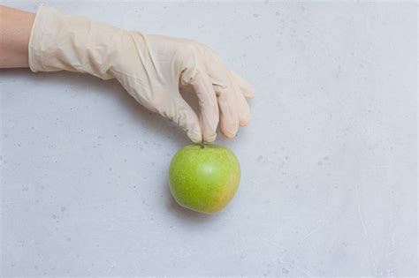 고무 장갑을 낀 손에는 녹색 사과 열매가 있습니다 녹색 사과는 밝은 회색과 흰색 현대 콘크리트 배경에 흰색 일회용 고무 라텍스