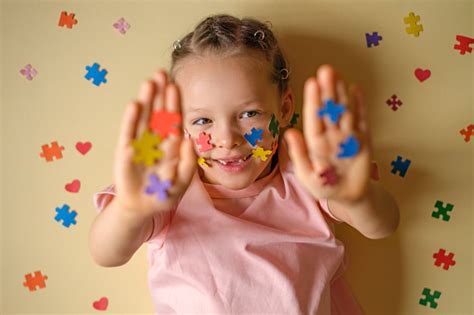 노란색 스튜디오 배경에 있는 아이는 여러 가지 빛깔의 작은 퍼즐 조각으로 장식되어 있습니다 소녀는 자폐증 상징이 있는 손바닥을 보여줍니다 지적 개발 및 교육 4 5세에 대한