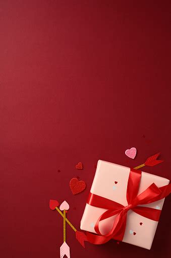빨간 리본이 달린 분홍색 종이로 싸인 발렌타인 데이 선물 상자는 하트 모양의 장식과 빨간색 배경에 큐피드의 화살표로 둘러싸여 있습니다 로맨틱하고 축제 테마에 적합합니다 2