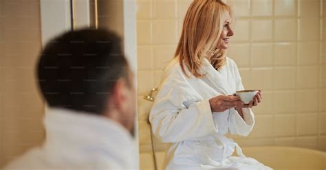 흰 가운을 입은 기쁜 금발 아가씨가 욕조 가장자리에 앉아 갈색 머리 남자친구와 차를 마시고 있다 사진 Unsplash의 공생 이미지