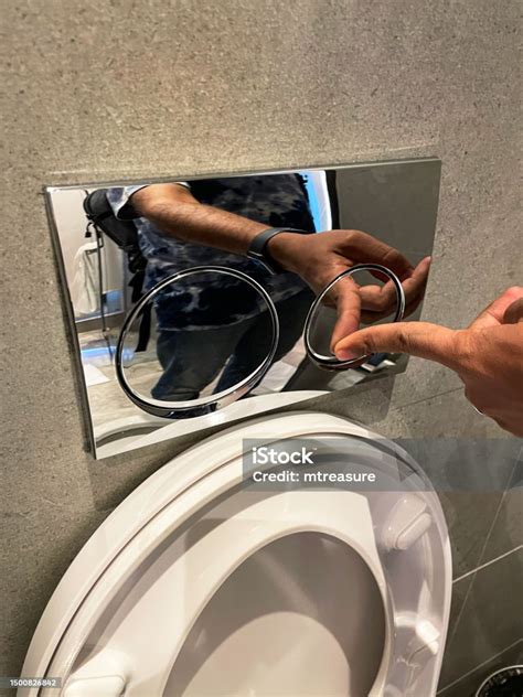 갈색 돌 타일 벽 벽걸이 형 반짝이는 크롬 이중 플러시 플레이트 흰색 세라믹 변기 뚜껑 변기 플러시 버튼을 누르는 인식 할 수없는 사람이 있는 고급 호텔 욕실의 클로즈업