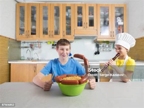 앞치마와 모자 요리사의 형태로 어린 소녀 미소 및 파이프 그리고 옆 앉아 어린 소년 연극과 배경 부엌에 뱀의 형태로 소시지와 함께 그릇에 보이는 2명에 대한 스톡 사진 및