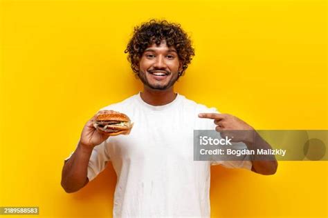 배고픈 남자 인도인이 맛있는 큰 햄버거를 들고 노란색 고립 된 배경에 표시 20 24세에 대한 스톡 사진 및 기타 이미지 20 24세 가리키기 건강에 좋지 않은 음식