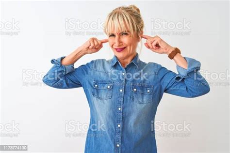 시끄러운 음악의 소음에 대한 성가신 표정으로 손가락으로 귀를 덮는 고립 된 흰색 배경 위에 서있는 캐주얼 한 데님 셔츠를 입은 중년 여성 청각 장애인 개념입니다 귀 부분에