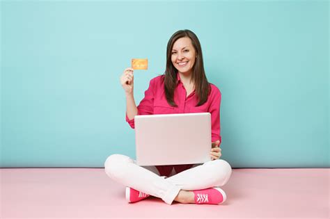 전체 길이 장미 셔츠에 젊은 여자의 초상화 바닥에 앉아 흰 바지 핑크 블루 파스텔 벽 배경 스튜디오에 고립 된 신용 카드 Pc 보유 패션 라이프 스타일 개념입니다 복사