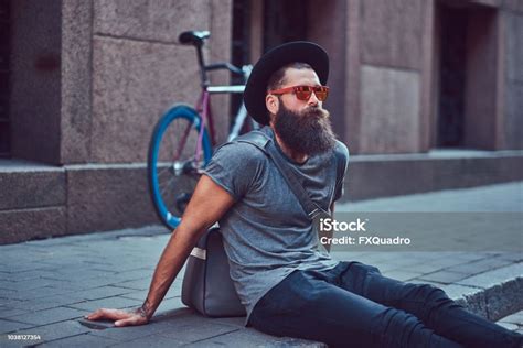 세련 된 수염과 가방 캐주얼 옷을 입은 그의 팔에 문신 한 잘생긴 Hipster 여행자 휴식 자전거를 타고 보도에 앉아 있다 T 셔츠에 대한 스톡 사진 및 기타 이미지