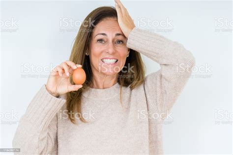 고립 된 배경 위에 신선한 계란을 들고 중 년 여성은 머리에 손으로 스트레스 수치심과 깜짝 얼굴이 충격 화가 좌절 두려움과 실수에 대 한 화가 갈색에 대한 스톡 사진 및