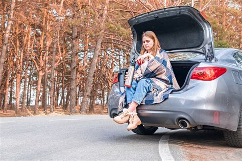 따뜻한 가을이나 봄날에 숲 옆에서 커피 컵과 보온병을 들고 자동차 트렁크에 앉아있는 젊은 여성 너 자신을위한 시간 묵상하고