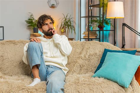 슬프고 외로운 인도 청년이 거실 소파에 앉아 인생 문제에 대해 생각하는 것처럼 보입니다 25 29세에 대한 스톡 사진 및 기타 이미지 25 29세 가정 생활 거실