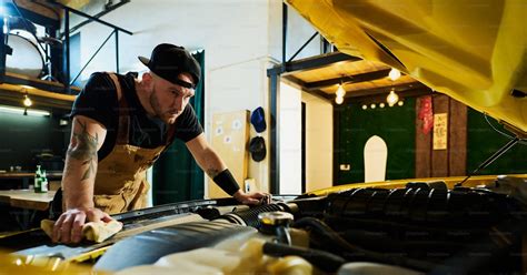앞치마와 야구 모자를 쓴 젊은 진지한 남자가 자동차의 열린 후드 위로 몸을 구부리고 차고나 작업장에서 엔진 점검을 하고 있다 사진 Unsplash의 자동차 이미지