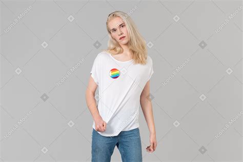 긴 금발 머리를 가진 잘 생긴 청년 회색 배경에 서서 청바지와 Lgbt 배지가있는 흰색 티셔츠를 입고 사진