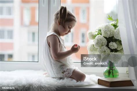 한 어린 소녀가 창턱에 앉아 있습니다 창가에 꽃병에 꽃다발과 꽃을 스니핑 소녀 창가에 흰색 꽃다발과 흰색 드레스에 작은 공주 가족에 대한 스톡 사진 및 기타 이미지 Istock