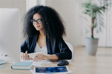 프리랜서 아프로 여성은 원격으로 작동 기쁘게 표현컴퓨터 화면에 초점을 맞춘 정보를 작성 넓은 캐비닛 배경에 화분에 공장에서 포즈 E 학습 개념 20 24세에 대한 스톡 사진