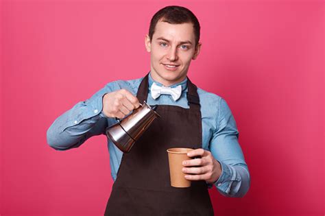 전문 남성 바리 스타의 초상화 종이 컵에 향기로운 커피를 붓는 파란색 셔츠를 착용 흰색 나비 넥타이와 갈색 앞치마 분홍색 배경에 고립 젊은 잘생긴 남자는 커피 숍에서 작동