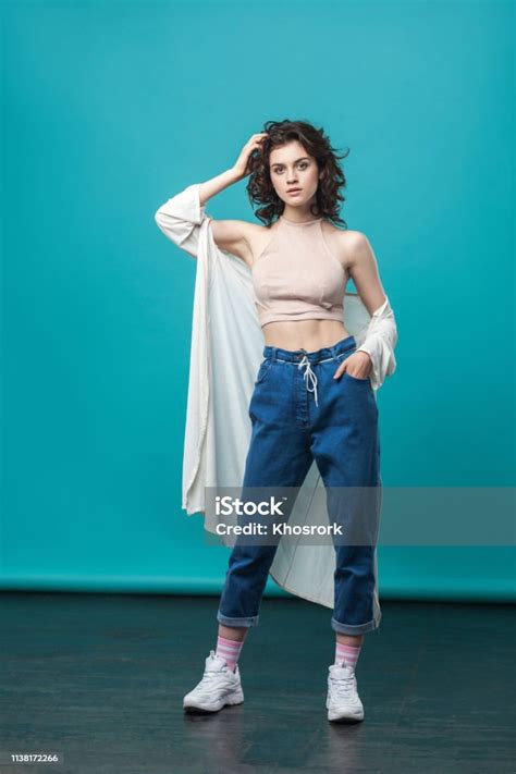 캐주얼 스타일 서 서와 푸른 벽에 대 한 포즈에 아름 다운 젊은 갈색 머리 여자의 전체 길이 심각한 얼굴로 카메라를 보고 가정의 방에 대한 스톡 사진 및 기타 이미지