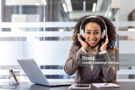 노트북이 있는 사무실에 앉아 흰색 헤드폰을 끼고 미소를 지으며 카메라를 보고 있는 아프리카계 미국인 젊은 여성 교육에 대한 스톡
