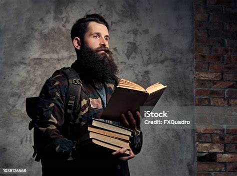 트렌디한 위장된 재킷을 입은 잘생긴 수염된 Hipster 어두운 배경에 고립 된 스튜디오에서 그의 손에 있는 책의 더미와 함께 서 있다 갈색 머리에 대한 스톡 사진 및 기타