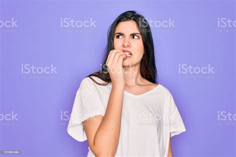 보라색 배경에 캐주얼 흰색 티셔츠를 입고 젊은 아름다운 갈색 머리 여자는 손톱을 물고 손톱을 물어 손에 스트레스와 긴장 찾고 불안 문제 물기에 대한 스톡 사진 및 기타