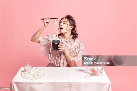 복고풍 스타일의 헤어도를 가진 젊은 아름다운 여성이 분홍색 배경에 고립 된 서빙 테이블에 앉아있는 중세 드레스를 입은 창조적 인
