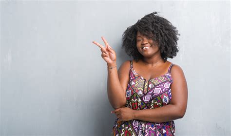 다채로운 입고 회색 그런 지 벽에 젊은 아프리카계 미국인 여자 드레스 승리 표시를 하 고 카메라에 윙크 하는 행복 한 얼굴로 웃 고 번호 2입니다 긍정적인 감정 표현에 대한