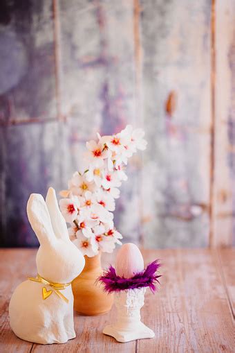 작은 흰색 기둥 위에 라일락 깃털이 있는 보라색으로 칠해진 부활절 달걀 아몬드 꽃의 가지 파란색 빈티지 배경에 흰색 부활절 토끼가 있고 복사 공간이 있습니다 달걀에 대한