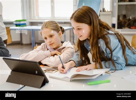 Curious Schoolgirls Doing E Learning Through Tablet PC Sitting At Desk In Classroom Stock Photo