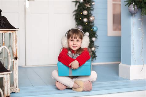 메리 크리스마스 행복한 휴일 새해 어린 소녀는 집 현관에 선물과 함께 빨간 스웨터에 앉아있다 어린이는 크리스마스를 위해 장식 된 테라스에 앉아 겨울 마당에서 재생됩니다