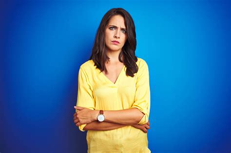 파란색 고립 된 배경 회의론자 와 긴장 위에 서있는 노란색 티셔츠를 입은 젊은 아름다운 여성은 교차 된 팔로 얼굴에 표정을