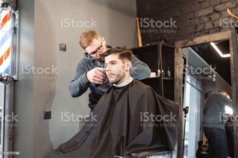 마스터는 이발소에서 남자의 머리와 수염을 잘라 미용사는 젊은 남자를위한 헤어 스타일을만든다 이발소에서 클리퍼 기계와 이발사 작업 2명에 대한 스톡 사진 및 기타 이미지