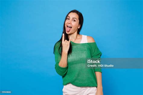 스튜디오의 밝은 파란색 벽 배경에 고립 된 중금속 바위 표지판을 묘사 한 혀를 보여주는 녹색 캐주얼 옷을 입은 쾌활한 젊은 여성의 초상화 사람 라이프 스타일 개념입니다 복사